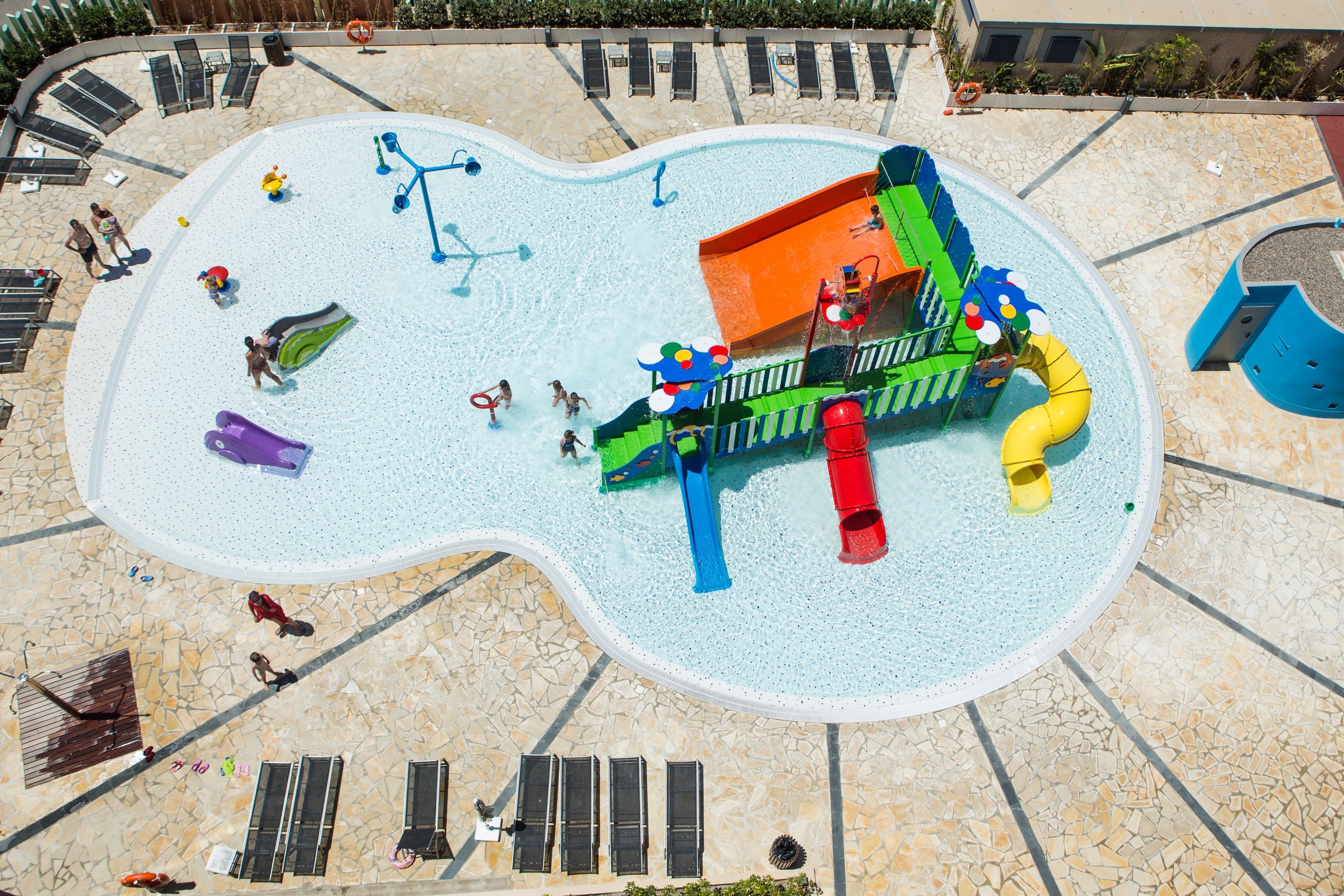 Uma grande piscina ao ar livre com diversas estruturas de recreação aquática, rodeada por espreguiçadeiras e um deck revestido de pedra.