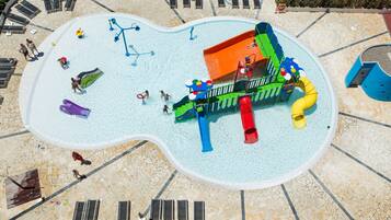 Uma grande piscina ao ar livre com diversas estruturas de recreação aquática, rodeada por espreguiçadeiras e um deck revestido de pedra.