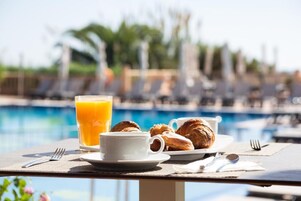 Mesa de café da manhã com croissants, uma xícara e um copo de suco de laranja à beira da piscina.