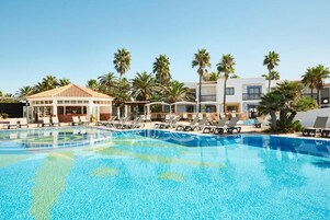Piscina com espreguiçadeiras, palmeiras e um edifício com telhado telhado.