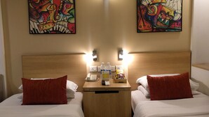 Quarto de hotel com duas camas, cada uma com travesseiros vermelhos, cabeceiras de madeira, uma mesinha com um telefone, uma garrafa de água e lanches. Há dois apliques de parede e duas pinturas abstratas coloridas acima das camas.