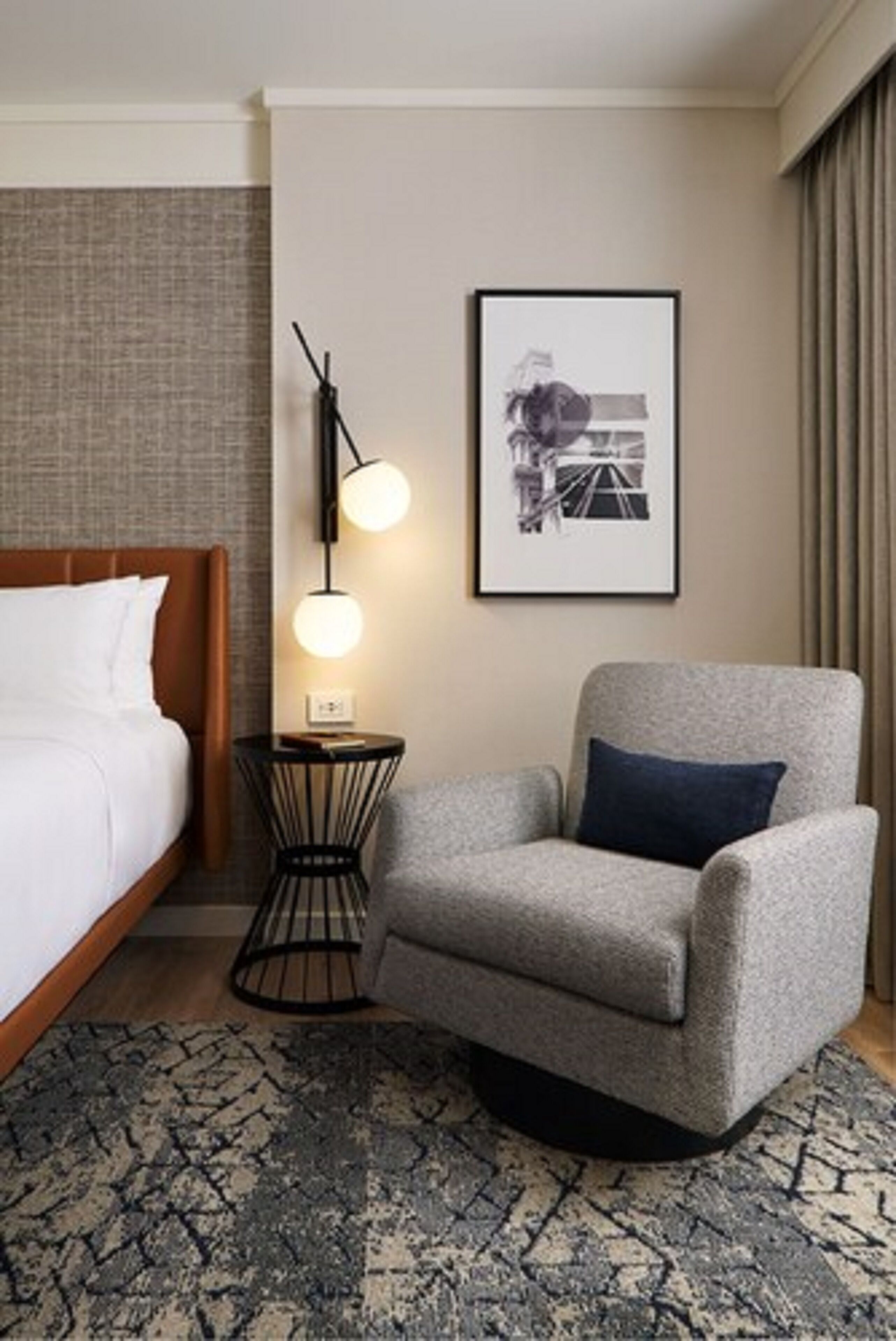 Quarto de hotel com uma cama, uma poltrona cinza, uma mesinha de cabeceira com uma luminária, um quadro na parede e um tapete estampado.