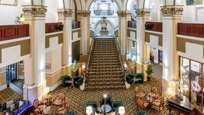 Um amplo hall de hotel com uma escadaria central, colunas ornamentadas e um candelabro.