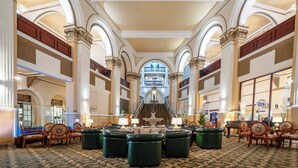 Um hall imponente com escadaria central, colunas decoradas e um lustre.