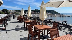 Um café ao ar livre com mesas e cadeiras de madeira, guarda-sóis bege e vista para o mar e a cidade ao fundo.