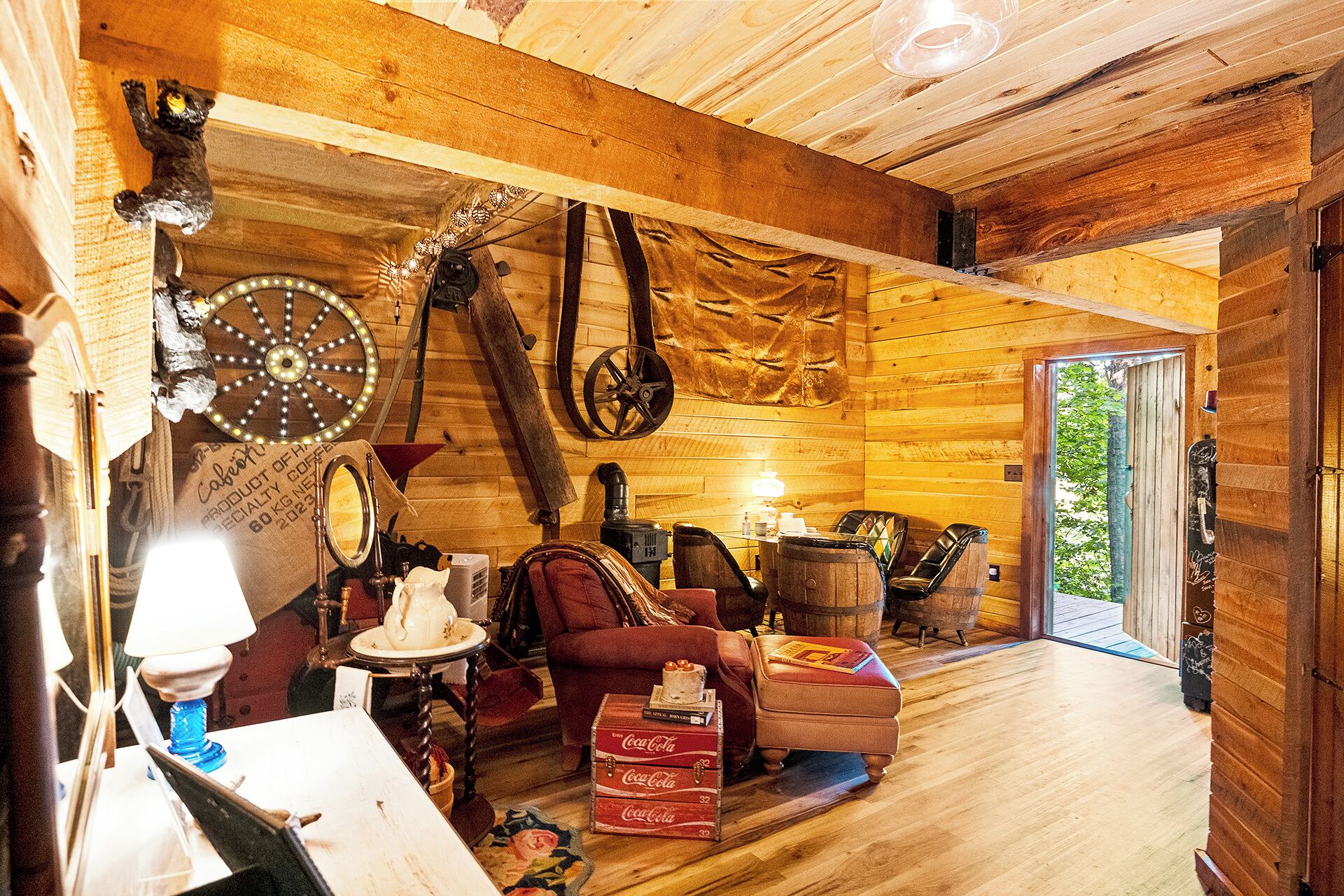Interior rústico com paredes e teto de madeira, uma grande roda, uma poltrona de couro, uma mesa pequena e uma porta que dá para o exterior.