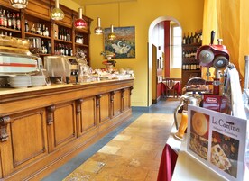 Interior de restaurante com balcão de madeira, prateleiras com garrafas, máquina de café e um aviso que diz 'La Cantina'.