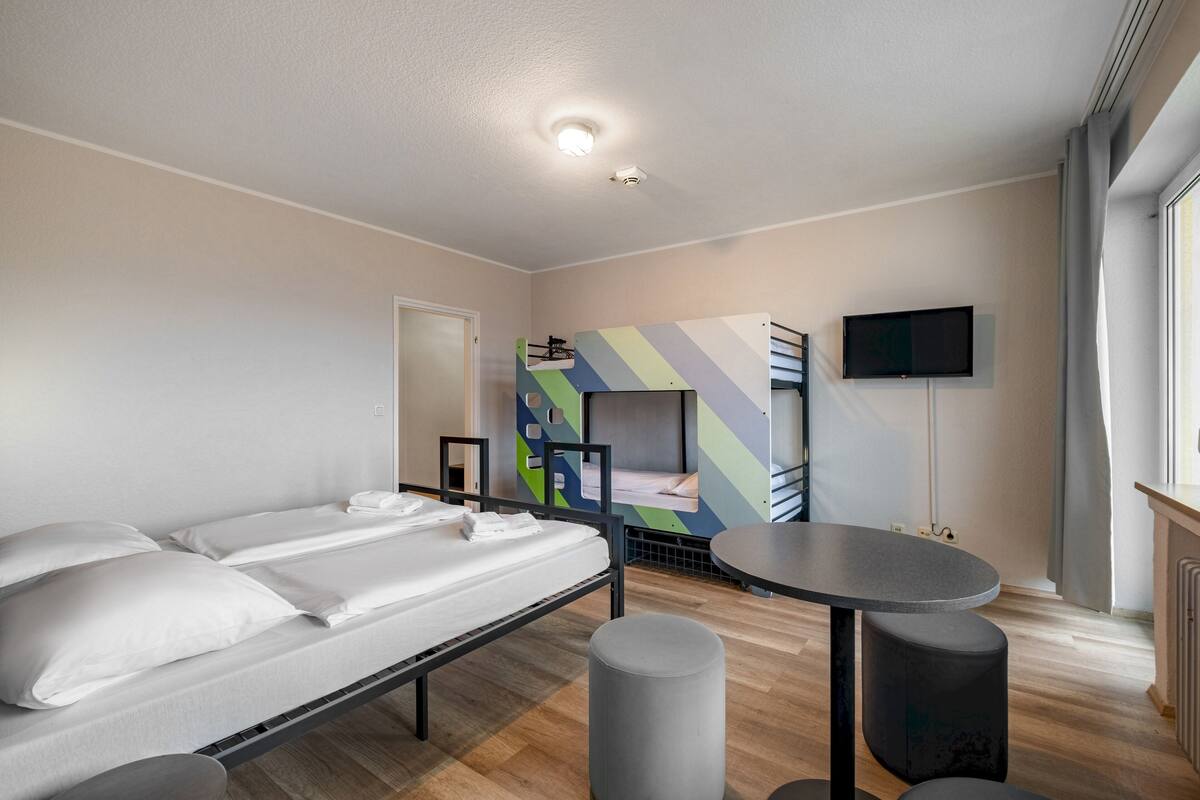 Quarto de hotel moderno com cama, uma mesinha, uma cadeira e televisão fixada na parede.