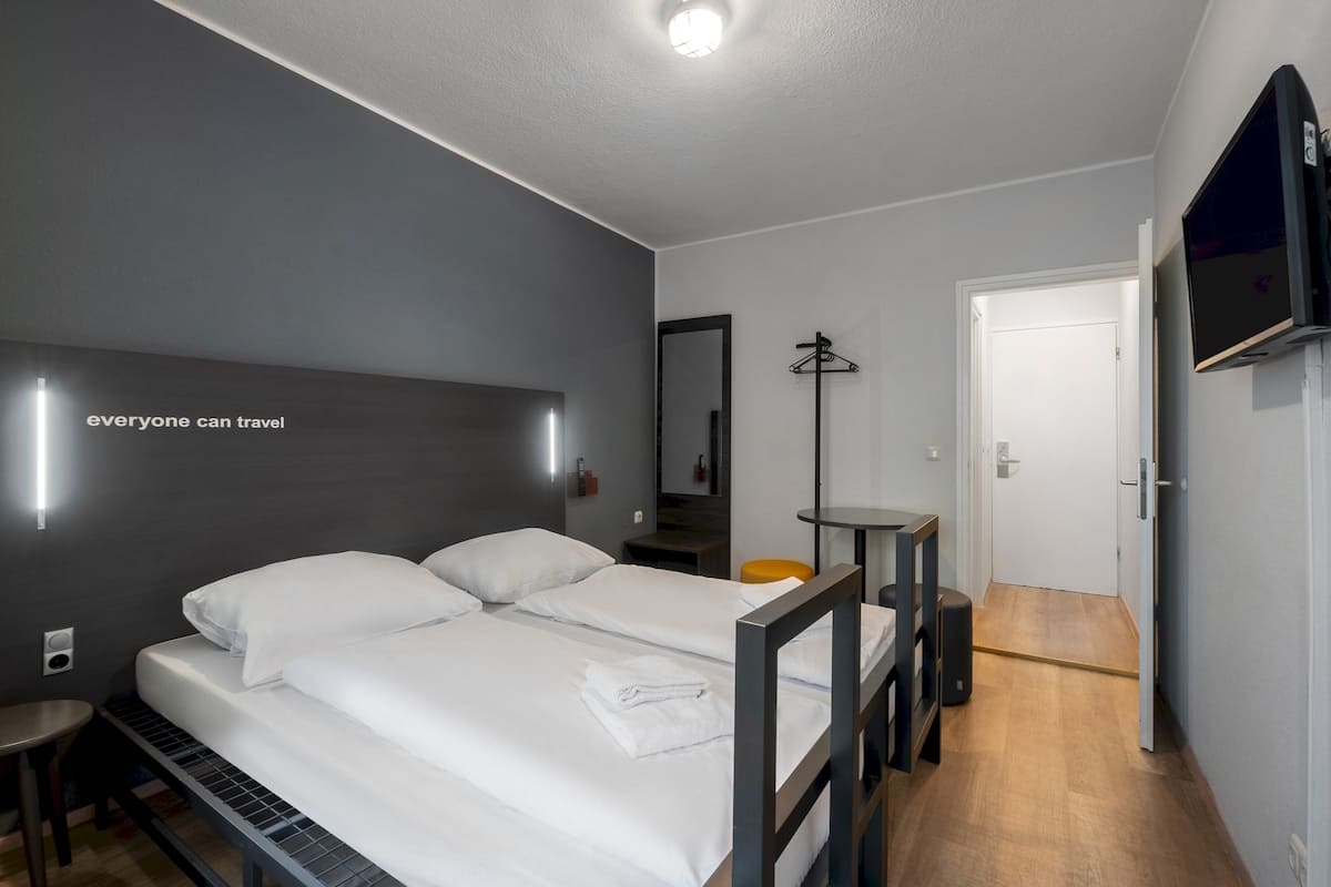 Quarto de hotel moderno com duas camas de solteiro, uma mesinha, um espelho, uma televisão e um porta-roupas.