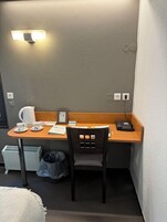Um pequeno espaço de escritório privativo com uma mesa, cadeira e telefone. Há uma cafeteira e uma lixeira.
