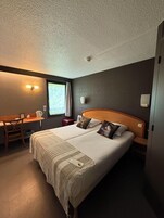 Quarto de hotel com uma cama grande, uma escrivaninha, uma cadeira, uma janela com cortinas e um ventilador de teto.