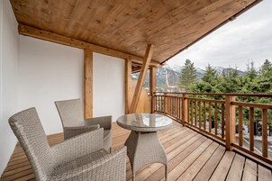 Um deck de madeira com cadeiras de vime e uma mesa de vidro, com vista para uma montanha nevada e uma floresta.