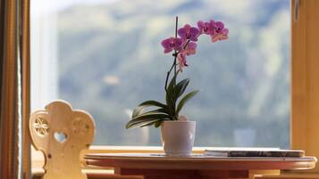 Uma orquídea em vaso sobre uma mesa de madeira, com um livro ao lado, perto de uma janela com vista desfocada para montanhas.