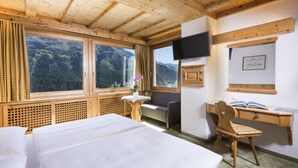 Quarto de madeira com uma cama, uma escrivaninha, uma cadeira, uma TV e uma janela com vista para as montanhas.
