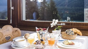 Mesa de café da manhã com café, frutas, doces e vista para montanhas pela janela.