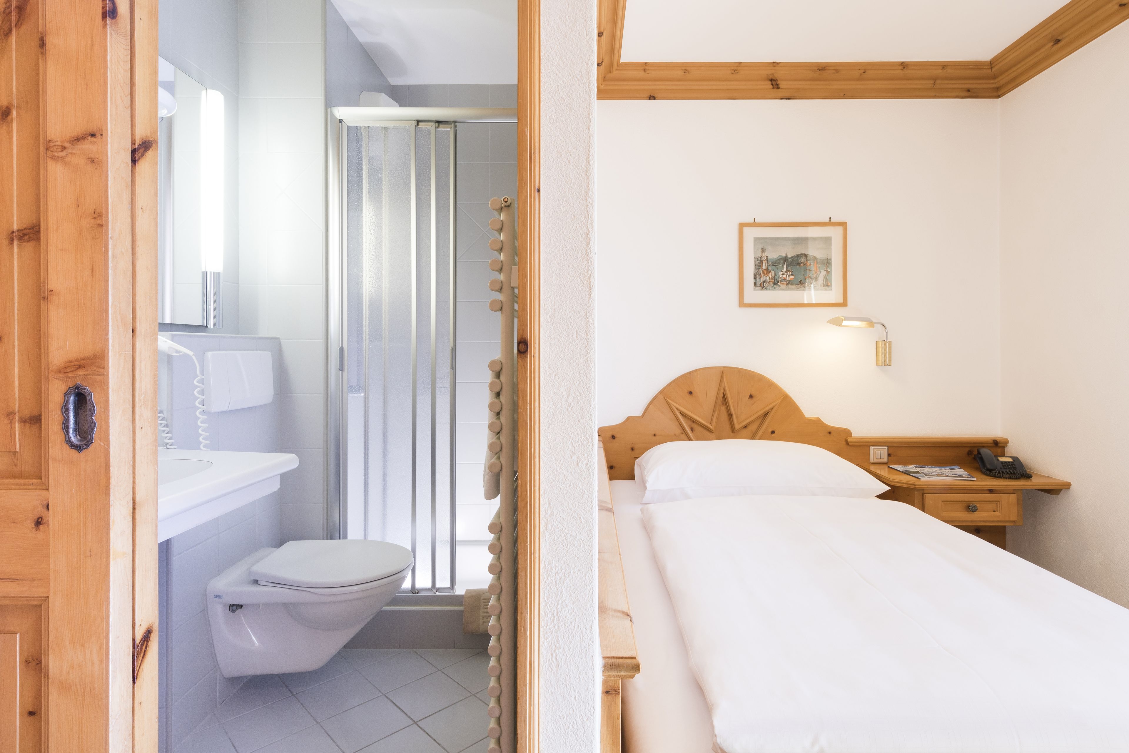 Quarto de hotel com cabeceira de madeira, mesa de cabeceira com luminária, quadro na parede e banheiro com vaso sanitário, pia e chuveiro.