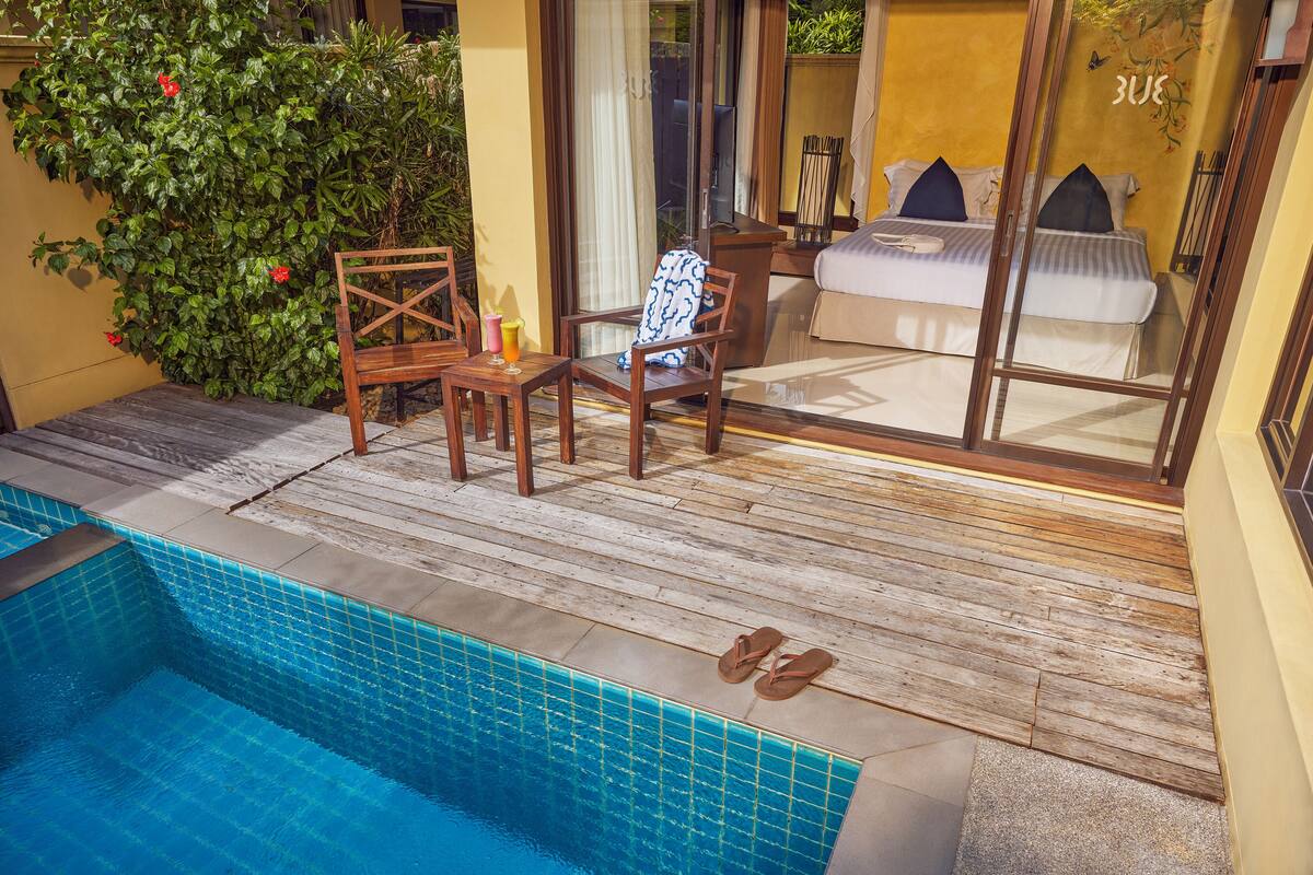 Uma área na piscina com um deck de madeira, uma cadeira, uma mesa com bebidas e uma cama visível através de uma porta de vidro.