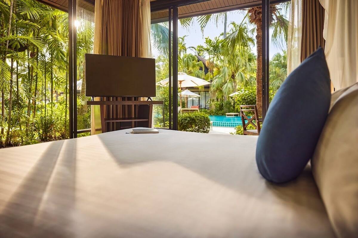 Quarto de hotel com uma cama grande, vista para uma piscina e vegetação exuberante lá fora.