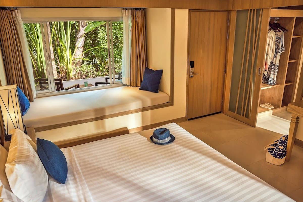 Quarto de hotel com cama, área de estar junto à janela, guarda-roupa e uma pequena mesa.