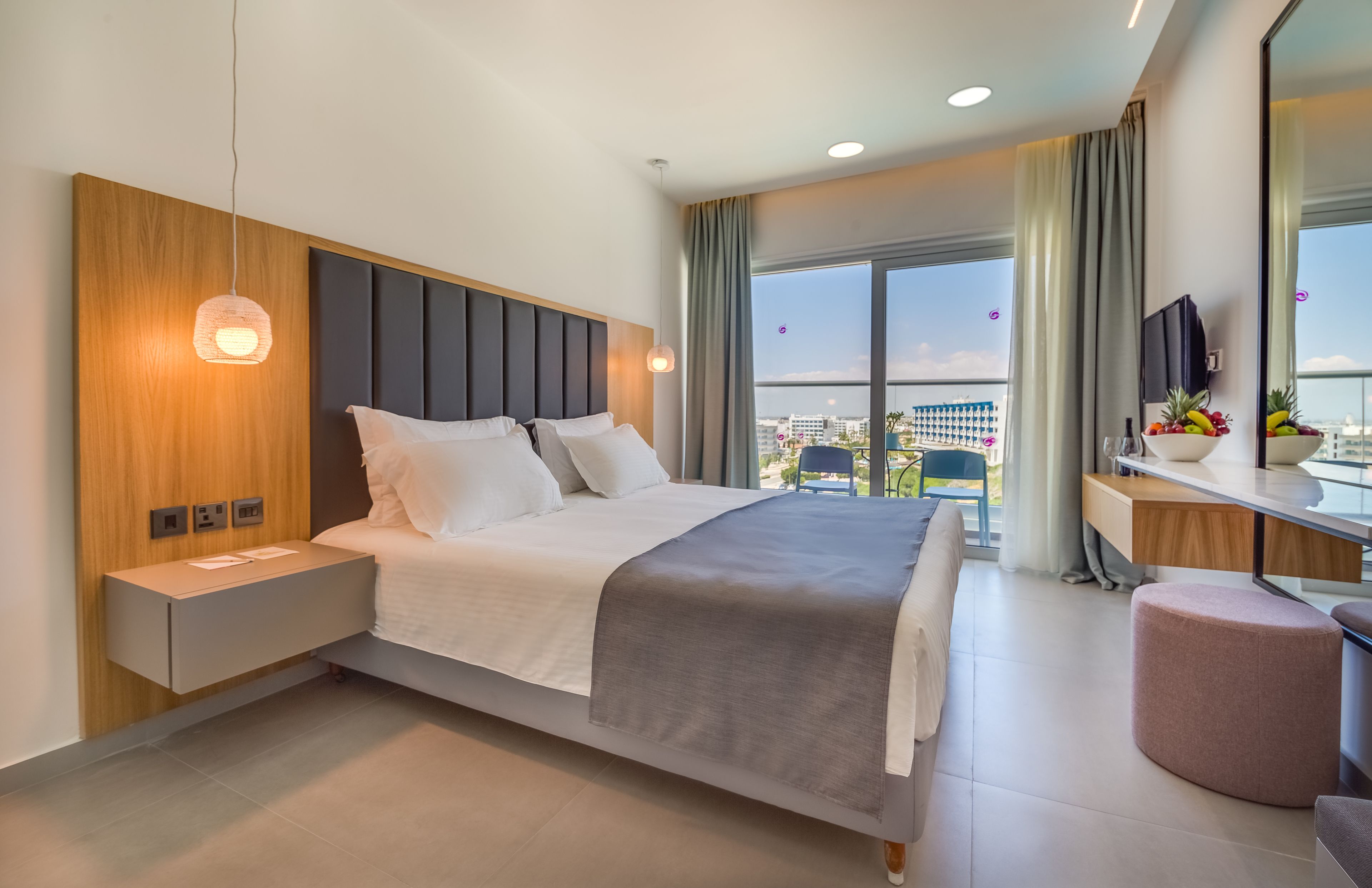 Quarto de hotel moderno com uma cama grande, mesas de cabeceira, uma escrivaninha e uma varanda com vista.