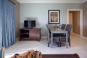 Uma sala de estar com televisão, mesa de jantar, cadeiras de vime e um quadro na parede.