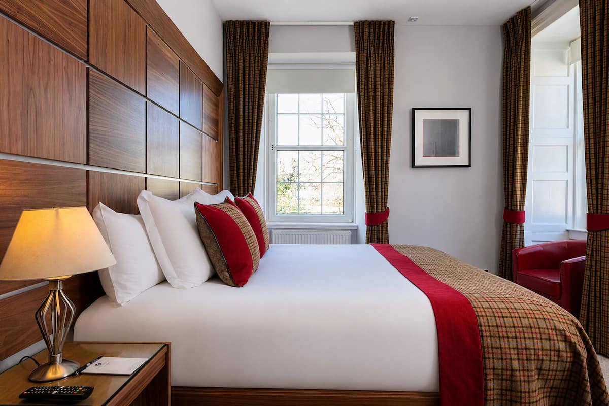 Quarto de hotel com uma cama grande, mesa de cabeceira com abajur, uma cadeira vermelha e uma janela com cortinas.