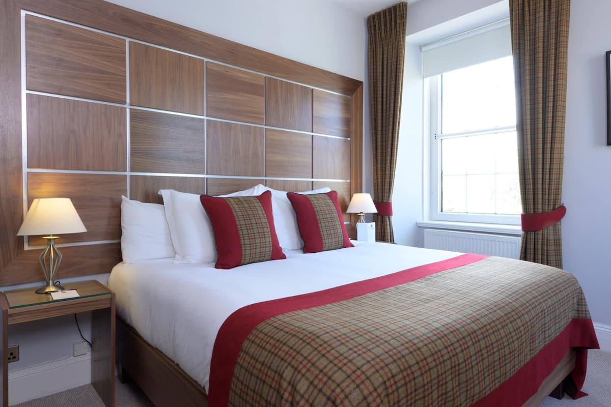 Cama bem arrumada com colcha xadrez e detalhes vermelhos, com duas mesas de cabeceira e luminárias, em um quarto com uma janela grande e uma cabeceira de madeira.