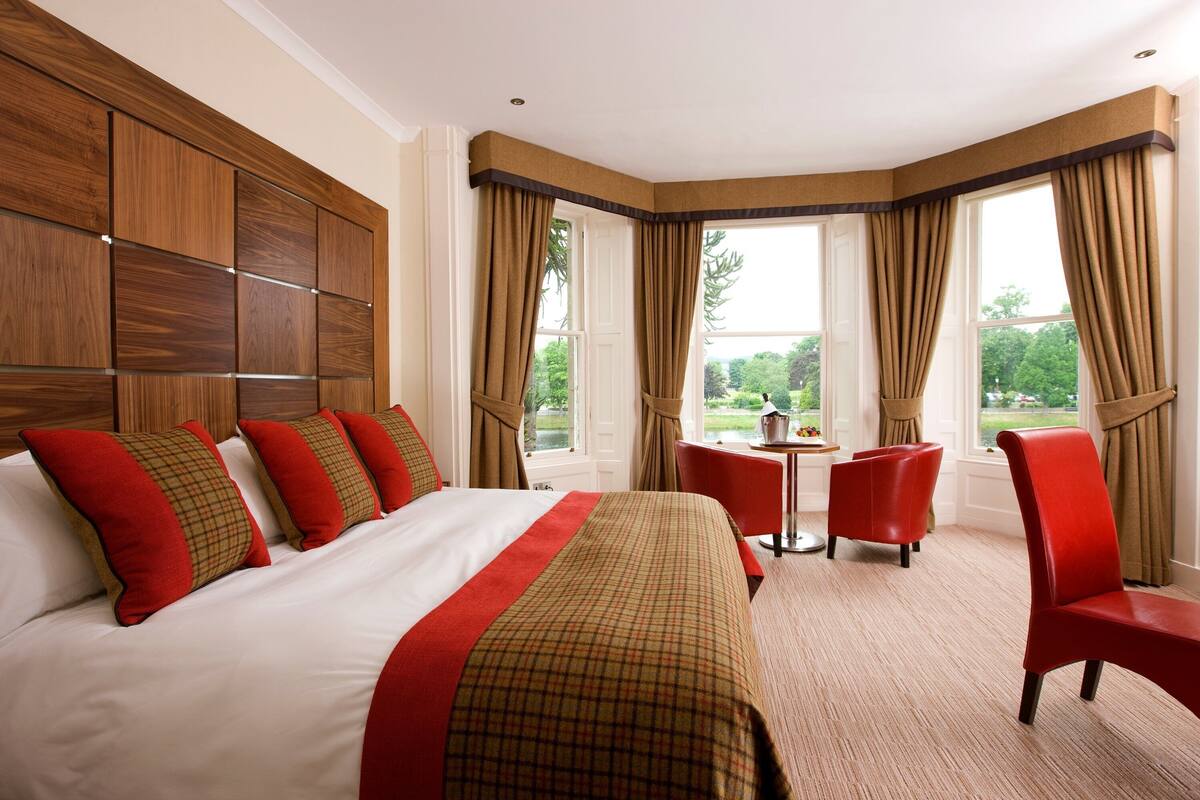 Quarto de hotel com uma cama grande, cadeiras vermelhas, uma mesinha e vista para o exterior.