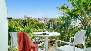 Uma varanda com cadeiras brancas, uma mesa com um livro e um vaso de vidro, e vista para edifícios e árvores.