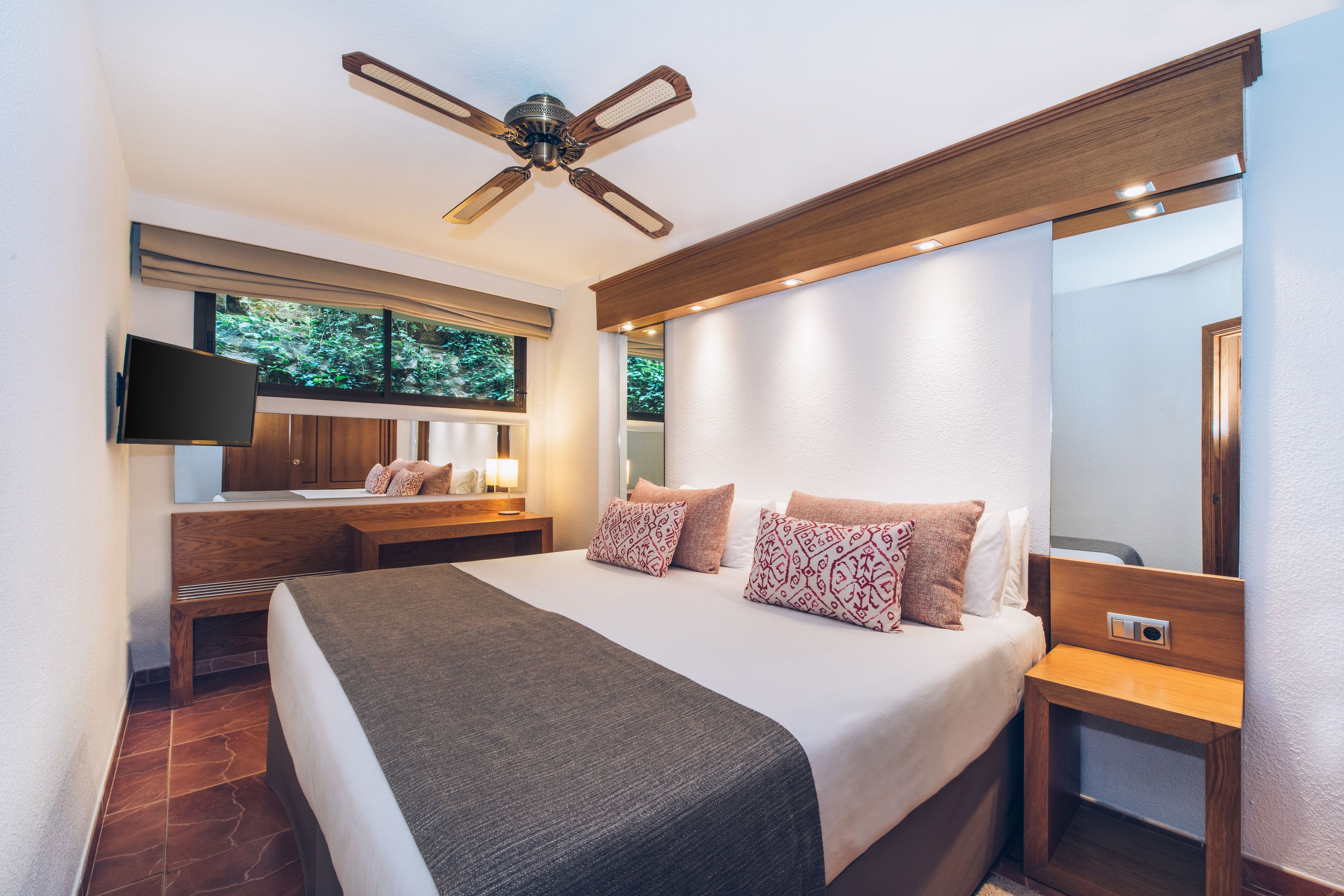Quarto de hotel moderno com uma cama grande, um ventilador de teto, uma televisão e uma janela com vista para árvores.