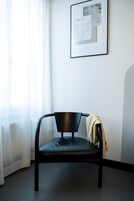 Uma cadeira com almofada escura e um casaco bege pendurado, colocada em um canto de um quarto com parede branca e quadro emoldurado acima.