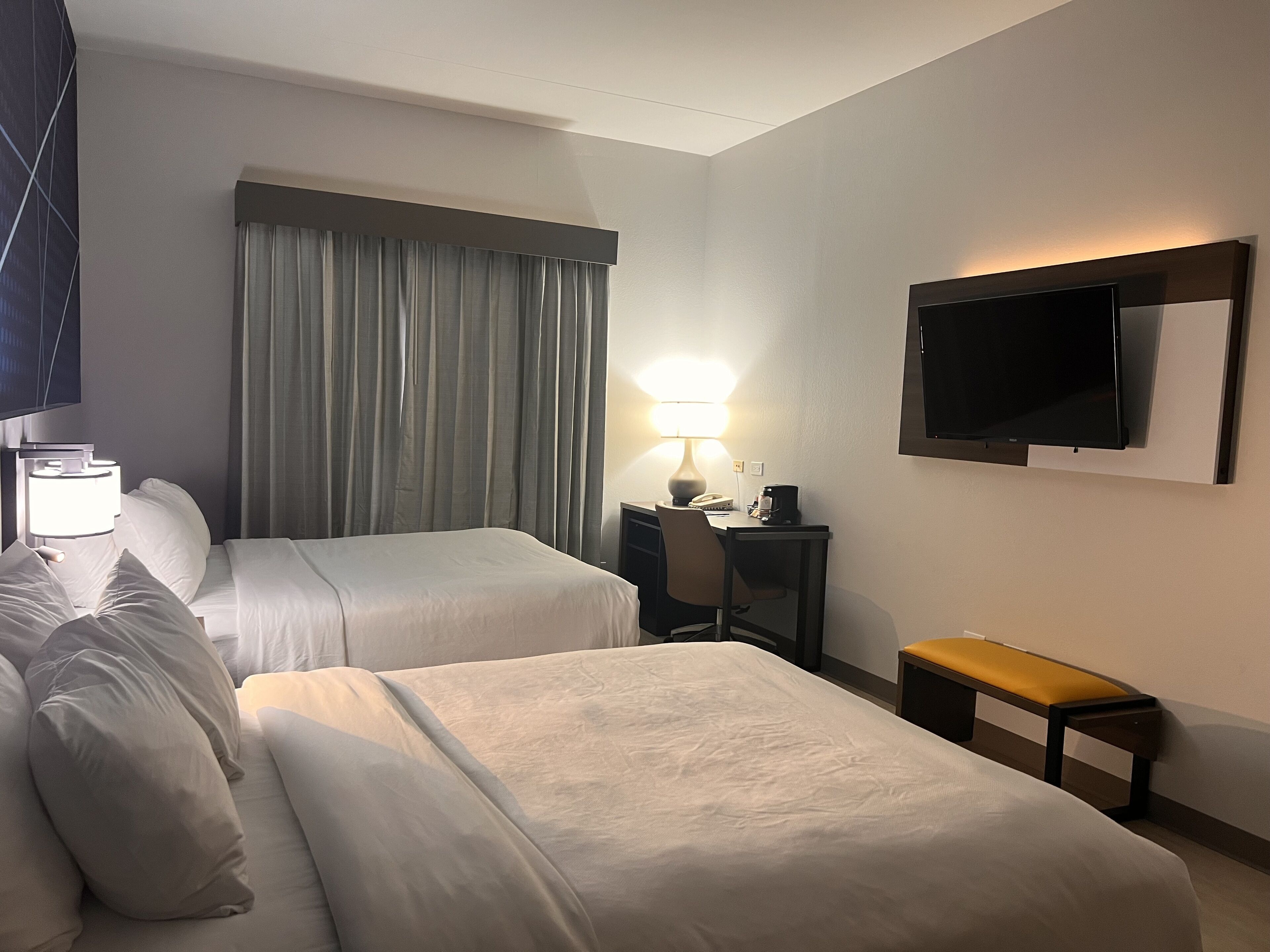 Quarto de hotel com duas camas, uma escrivaninha com luminária, televisão e um pequeno banco.