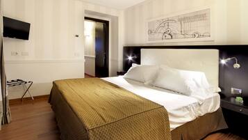 Quarto de hotel com uma cama, televisão fixada na parede, uma obra de arte emoldurada acima da cama e uma porta que dá para outro cômodo.