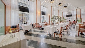 Um restaurante espaçoso com tetos altos, grandes janelas e chão xadrez. As mesas estão dispostas com toalhas brancas e cadeiras.