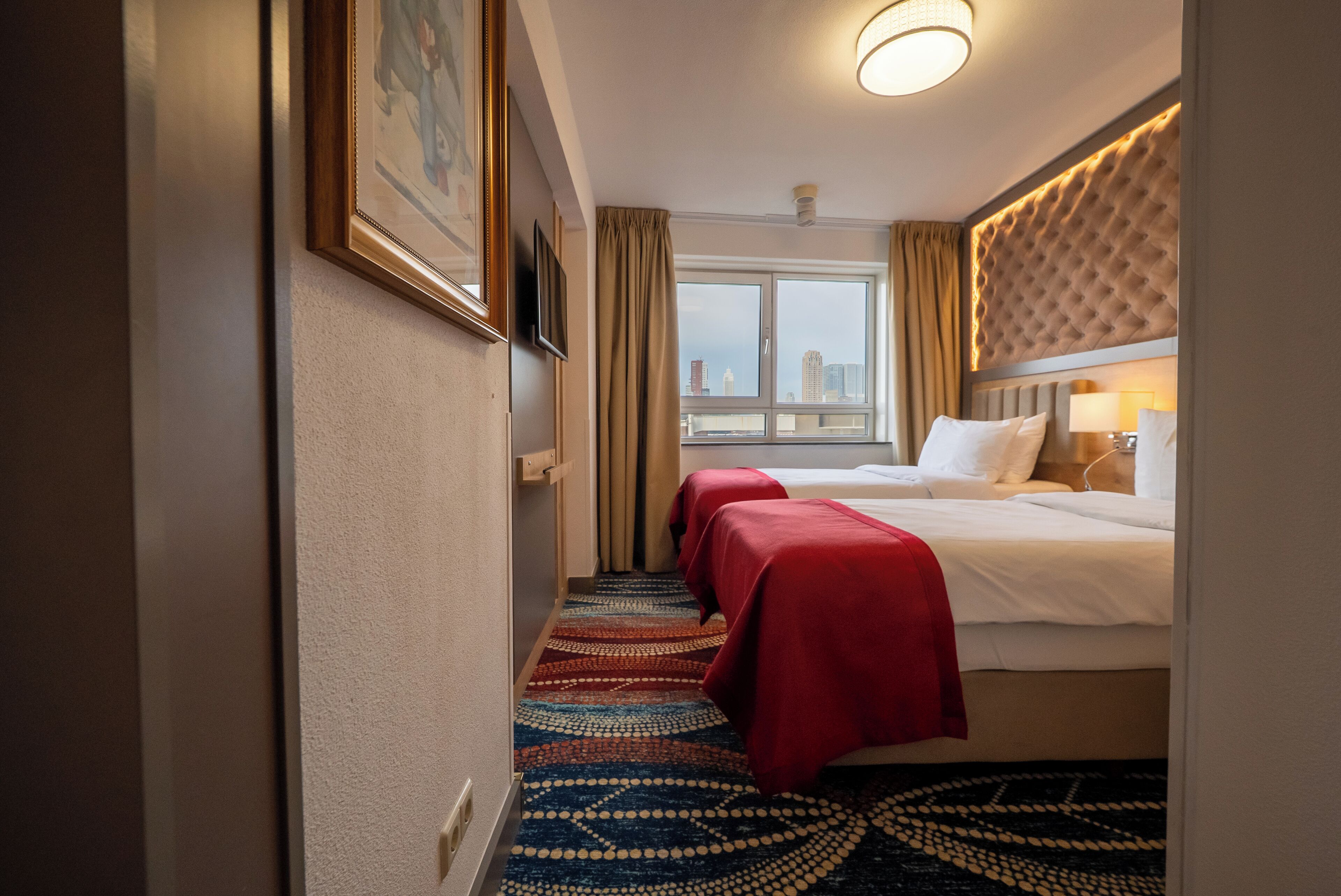 Quarto de hotel com duas camas, uma janela grande com vista para a cidade, um tapete estampado e um cobertor vermelho sobre uma das camas.