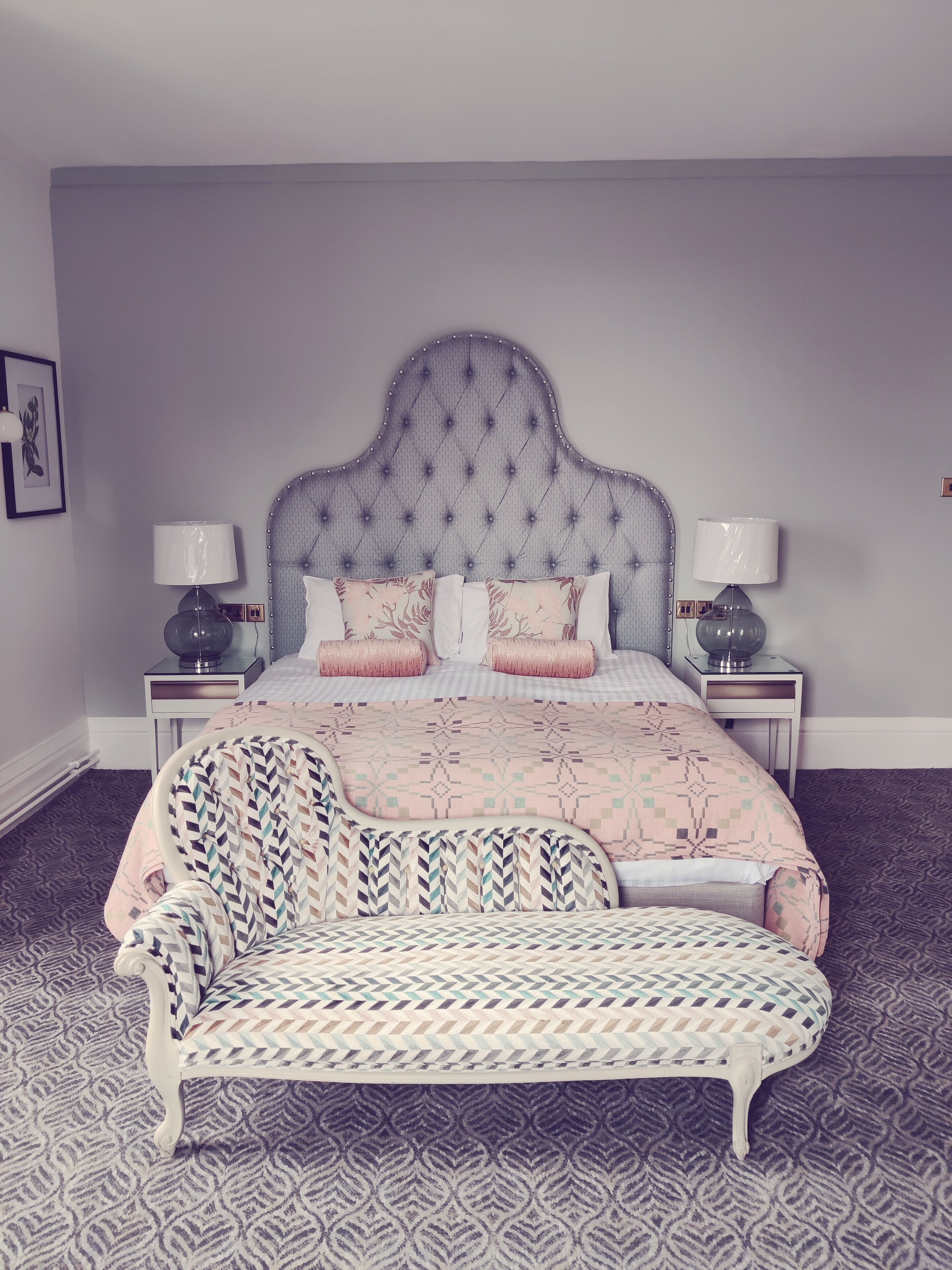 Quarto com cabeceira estofada, colcha estampada, duas luminárias de cabeceira e um sofá estampado.