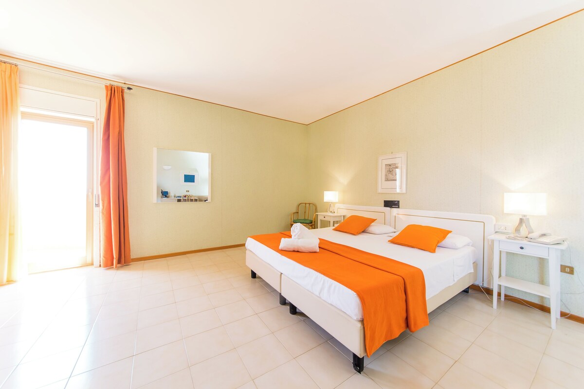Quarto de hotel com uma cama grande, duas mesas de cabeceira, um criado-mudo, um espelho e uma janela com cortinas.
