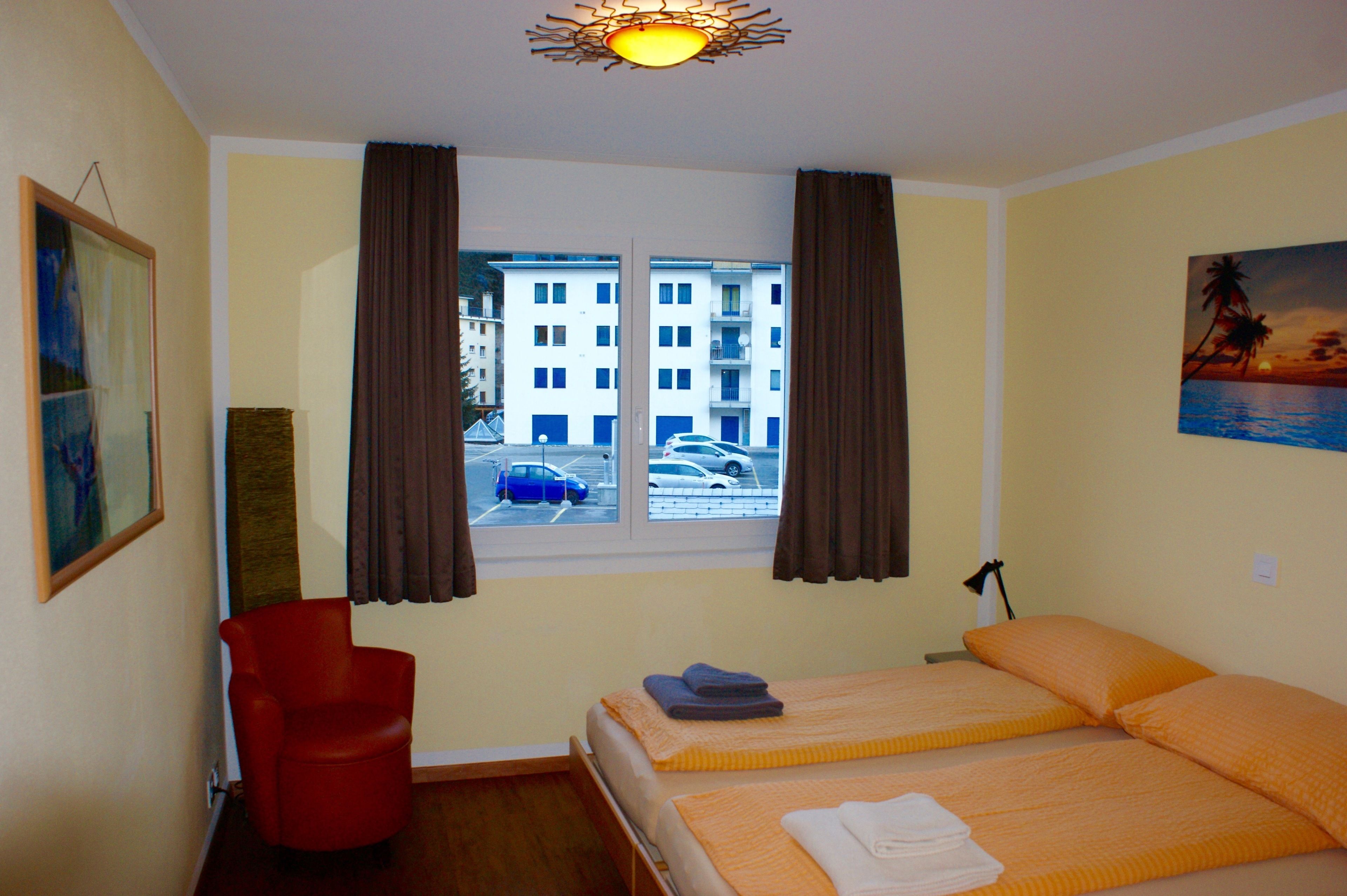 Um quarto com uma cama, uma cadeira, uma janela com cortinas, um quadro na parede e um lustre no teto.