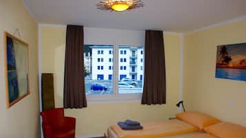 Um quarto com uma cama, uma cadeira, uma janela com cortinas, um quadro na parede e um lustre no teto.