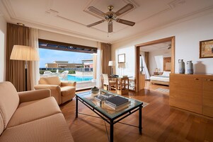 Quarto de hotel com mesa de centro de vidro, poltronas bege, um armário de madeira e vista para uma piscina.