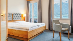 Quarto de hotel com cama de madeira, uma cadeira, varanda com vista para montanhas e janela com cortinas.
