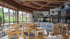Um restaurante com mesas e cadeiras de madeira, janelas amplas e uma área de cozinha.