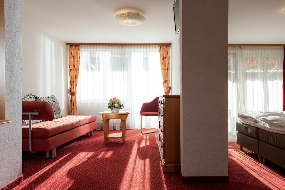 Quarto de hotel com sofá, uma pequena mesa com um vaso de flores, uma cadeira, cômoda e cama.