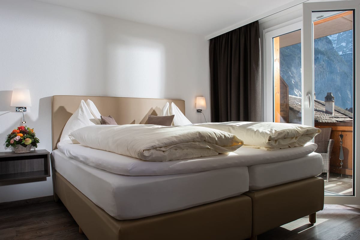 Quarto de hotel moderno com uma cama grande, duas mesas de cabeceira com luminárias, uma pequena planta e vista para uma montanha através de uma porta deslizante.