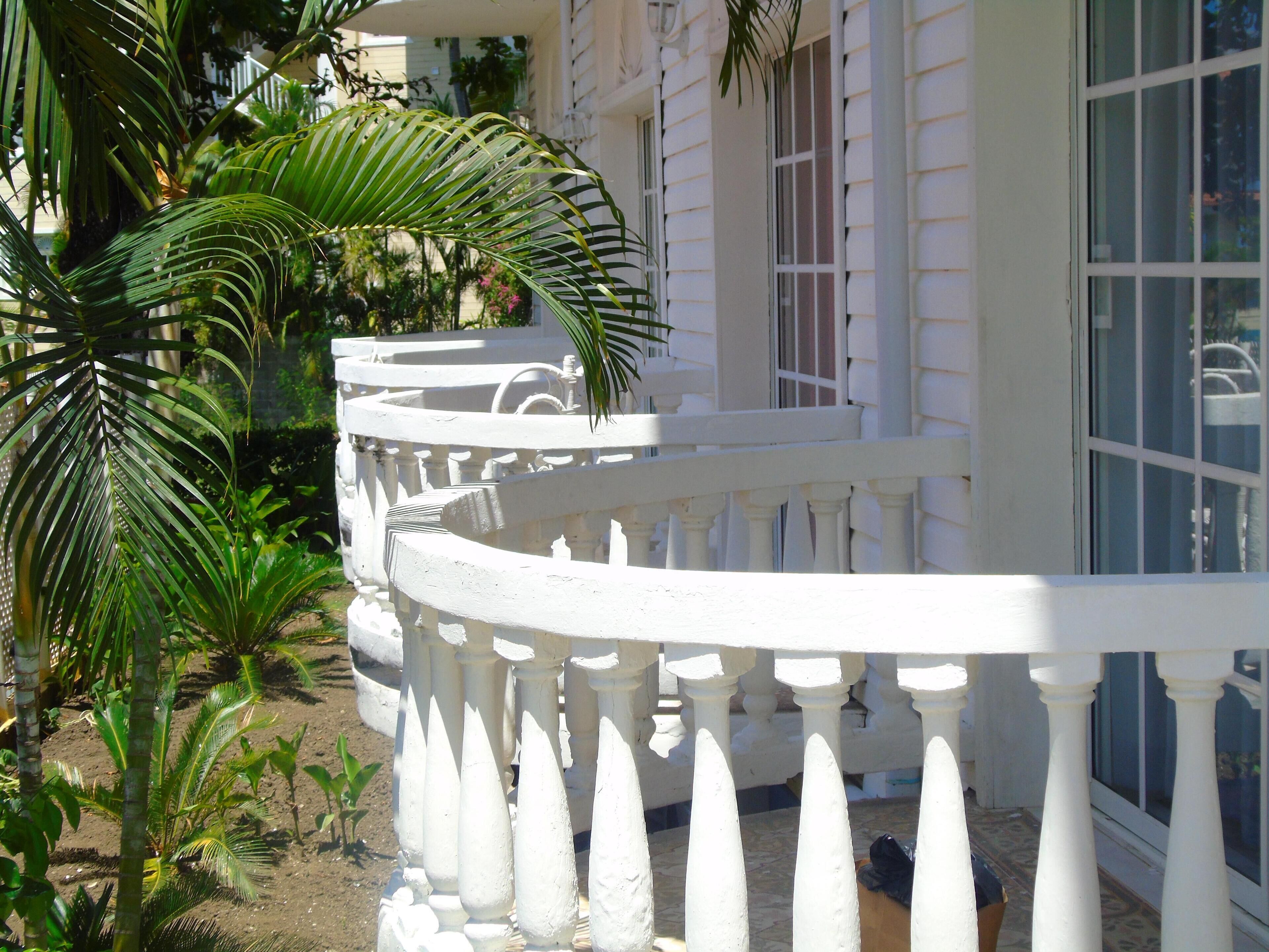 Uma varanda branca com corrimão curvo, rodeada por plantas verdes exuberantes e um edifício com janelas amplas.