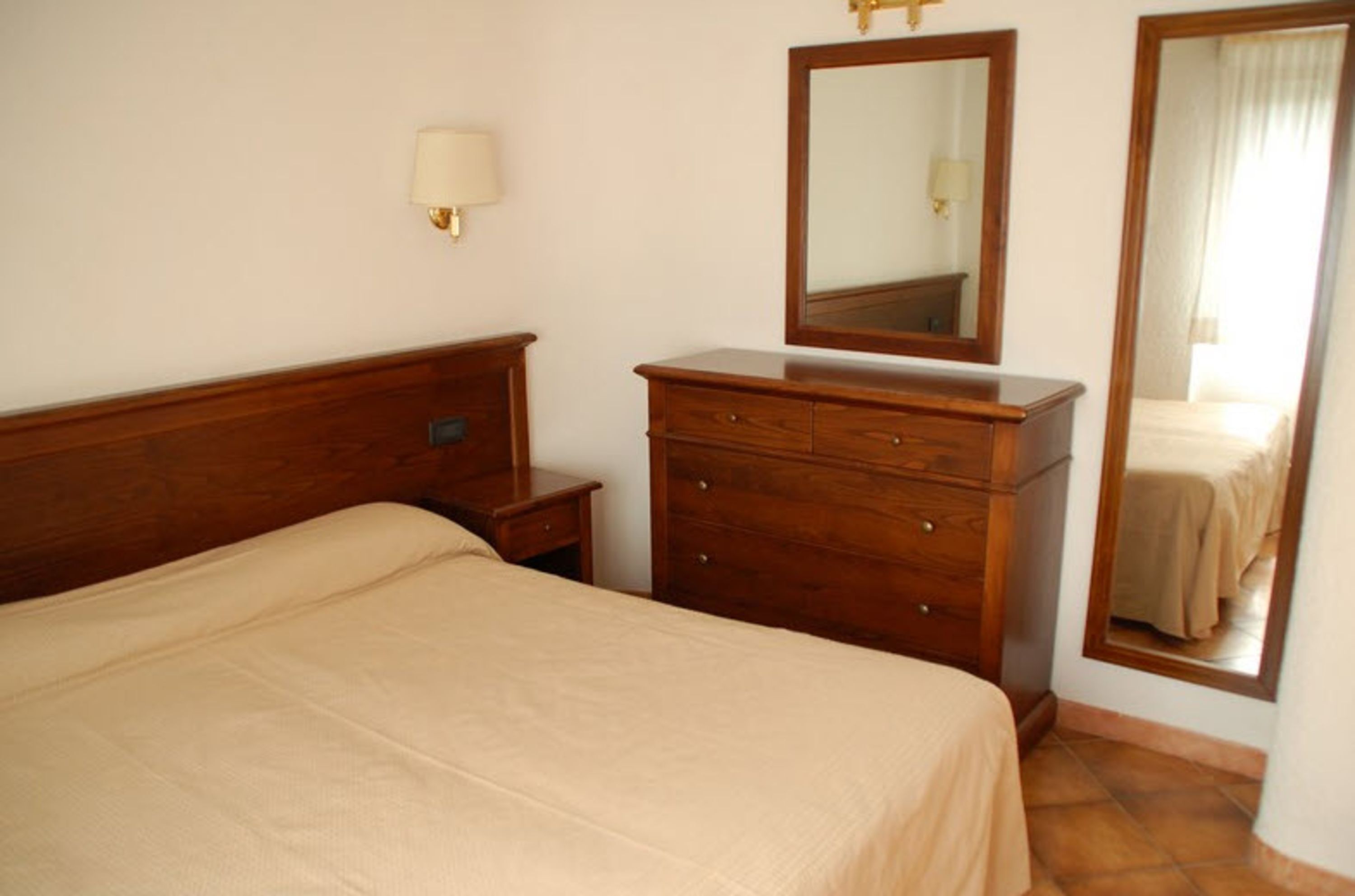 Quarto com cama de madeira, cômoda, mesa de cabeceira, luminária na parede e espelho.