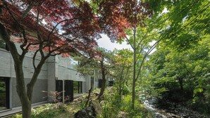 Um edifício moderno com fachada de vidro, cercado por uma paisagem natural com árvores e um riacho.