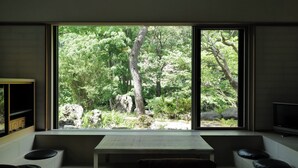 Quarto moderno com uma grande janela com vista para uma floresta. Há uma mesa de madeira e um banco com almofadas.