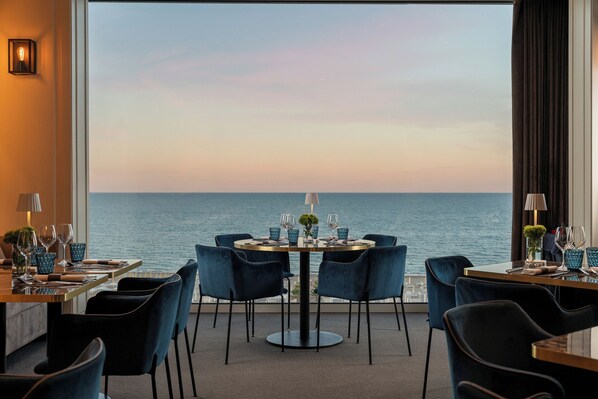 Uma área de refeições com uma mesa posta para quatro pessoas, de frente para uma grande janela com vista para o mar.