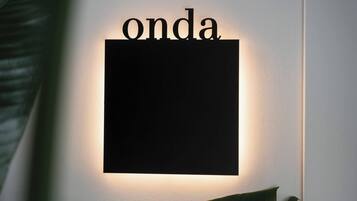 Obra emoldurada com a palavra "onda" em uma parede, rodeada por folhagem verde.